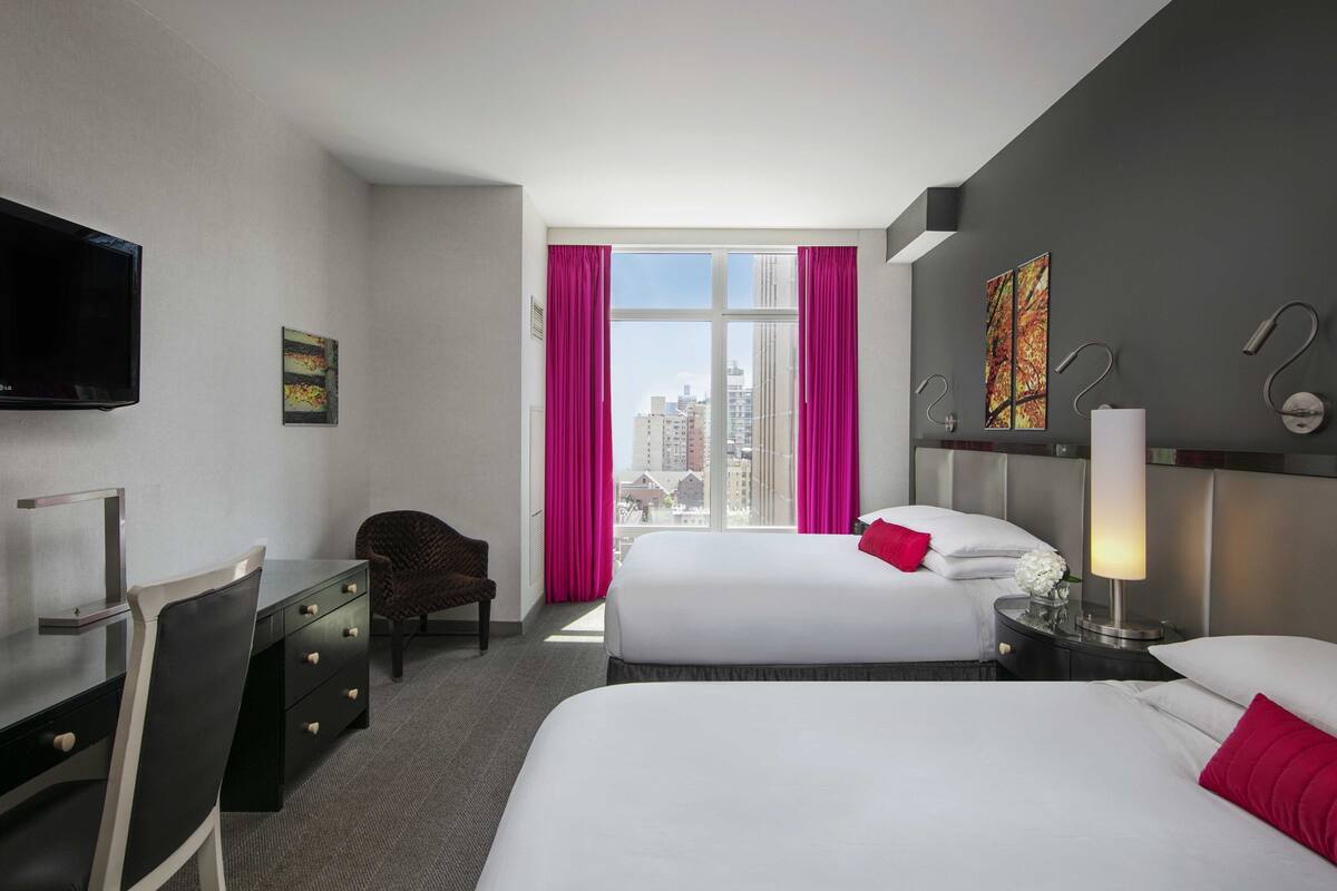 Quarto de hotel com duas camas, uma mesa, uma cadeira, uma televisão e uma janela com cortinas.