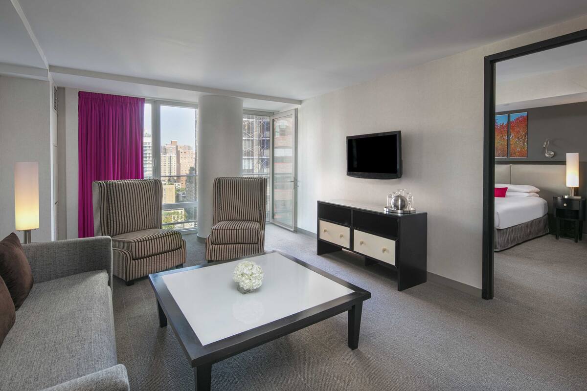 Quarto de hotel com sofá, duas poltronas listradas, mesa de centro, televisão, mesa de cabeceira com abajur, cama e vista da cidade por grandes janelas.