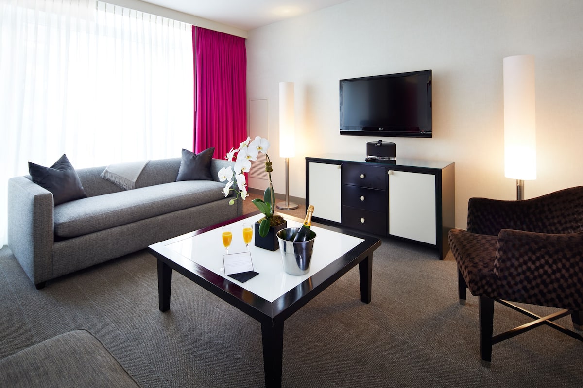 Quarto de hotel moderno com sofá cinzento, poltrona preta, mesa de centro branca com uma garrafa de champanhe, copos e um arranjo floral. Uma televisão de tela plana está fixada na parede e há duas luminárias de chão.
