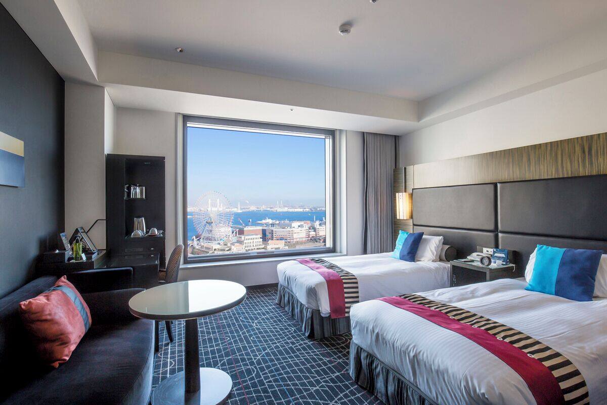 Quarto de hotel com duas camas, um sofá, uma mesa pequena, uma TV e uma janela grande com vista para a cidade.