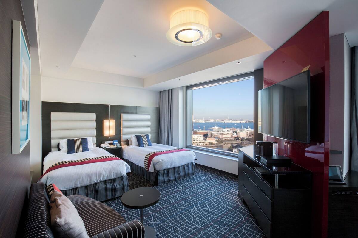 Quarto de hotel com duas camas, um sofá, uma TV e uma janela grande com vista para a cidade.