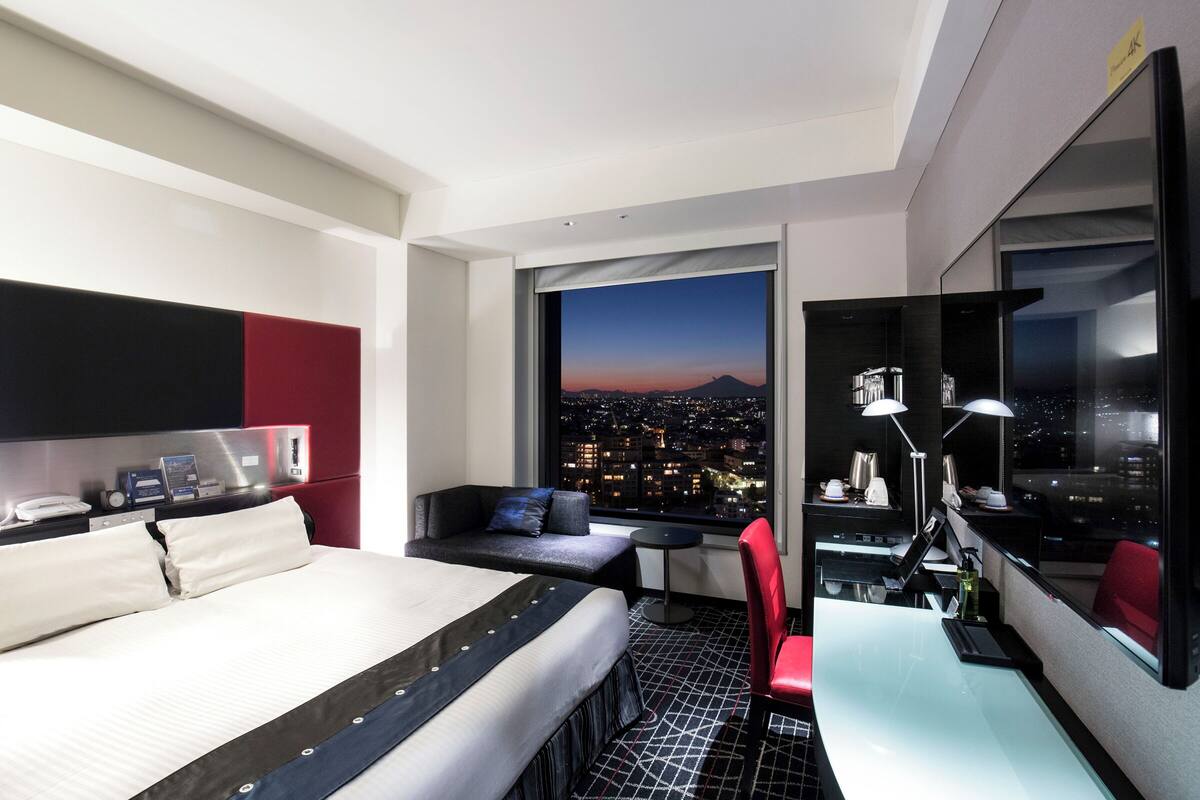 Quarto de hotel com cama, escrivaninha, cadeira, sofá, uma janela grande com vista para a cidade e uma televisão.