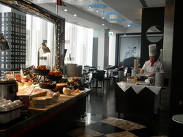 Restaurante com buffet, cozinheiro preparando comida e uma grande janela com vista para a cidade.
