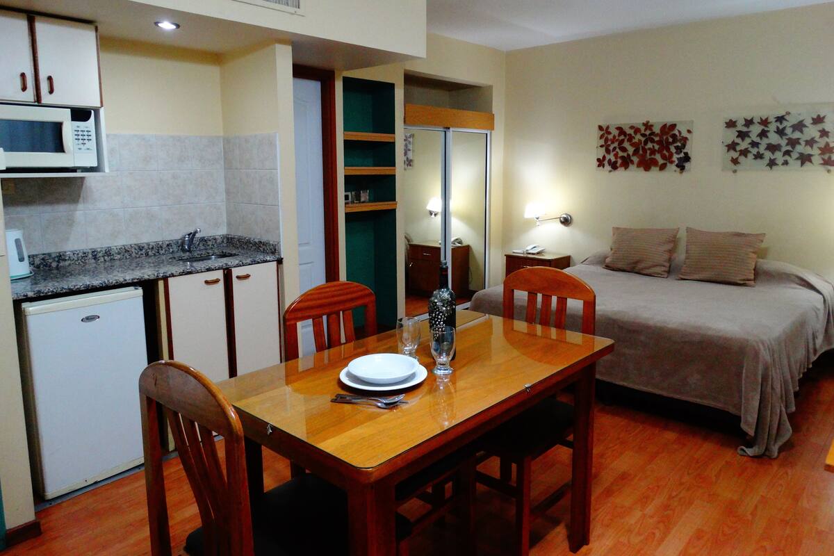 Quarto compacto com cozinha compacta, uma mesa de jantar com cadeiras, uma cama e um espelho.