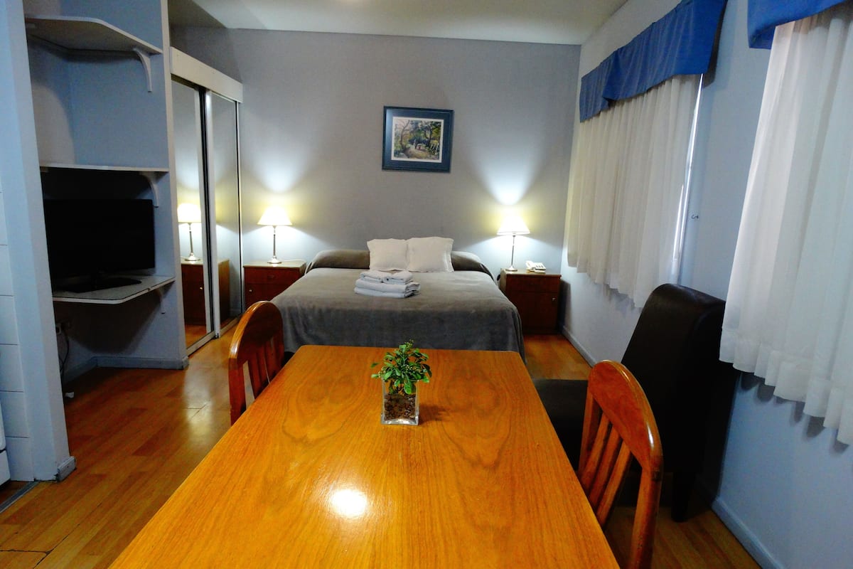 Quarto de hotel com mesa de jantar de madeira, cama com colcha cinza, duas luminárias de cabeceira, um quadro na parede e uma televisão sobre um suporte.