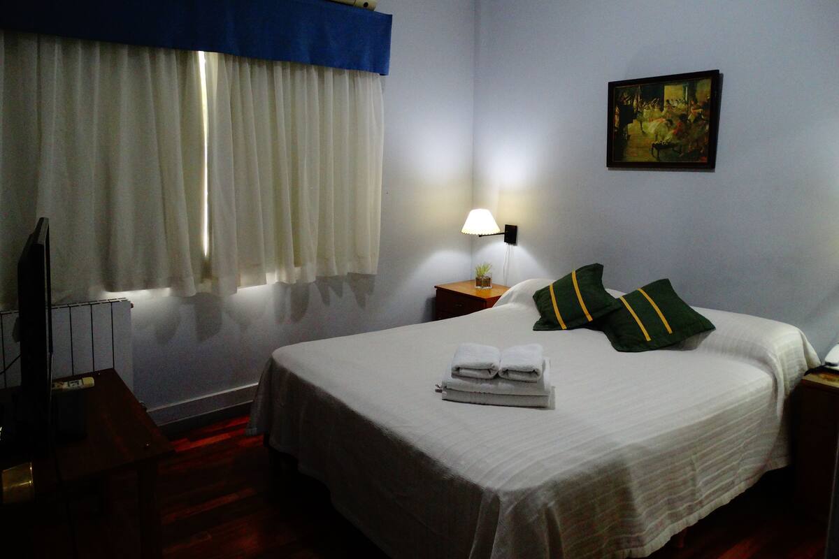 Quarto de hotel com cama, mesa de cabeceira, luminária, um quadro na parede e ar condicionado sobre a cama.