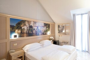 Quarto de hotel com uma cama grande, mesa de cabeceira com telefone, espelho e uma pintura de uma paisagem costeira.
