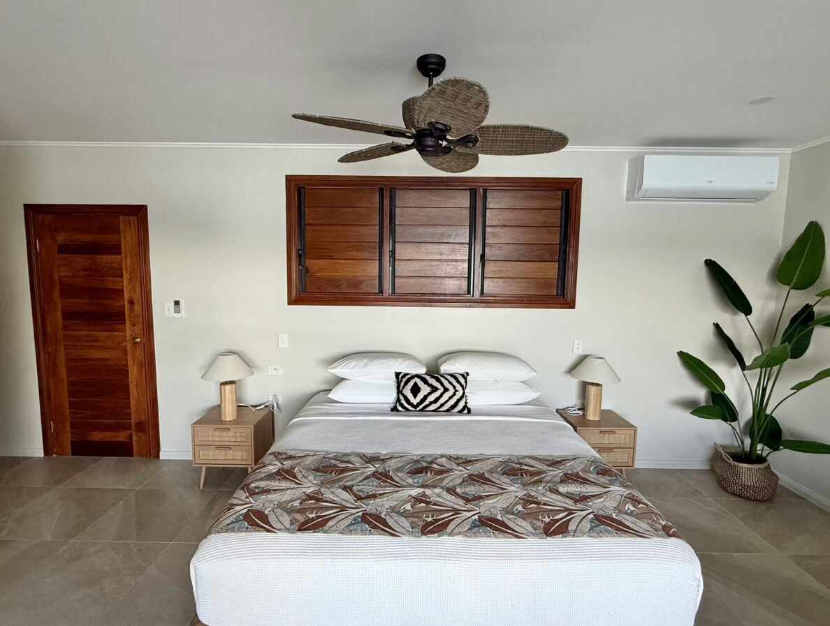 Quarto com cama, cabeceira de madeira, ventilador de teto, abajures e uma planta em vaso.