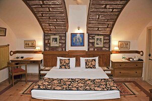Quarto com uma cama grande, cabeceira de madeira e teto de pedra. Há duas mesas de cabeceira com luminárias, uma escrivaninha e uma cadeira.
