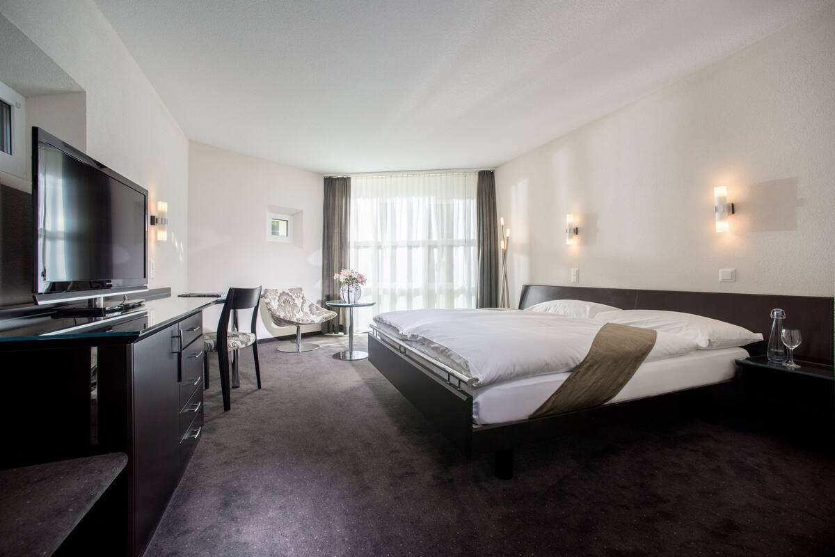 Quarto de hotel com cama, escrivaninha com televisão, cadeira, mesinha com um vaso e janela com cortinas.