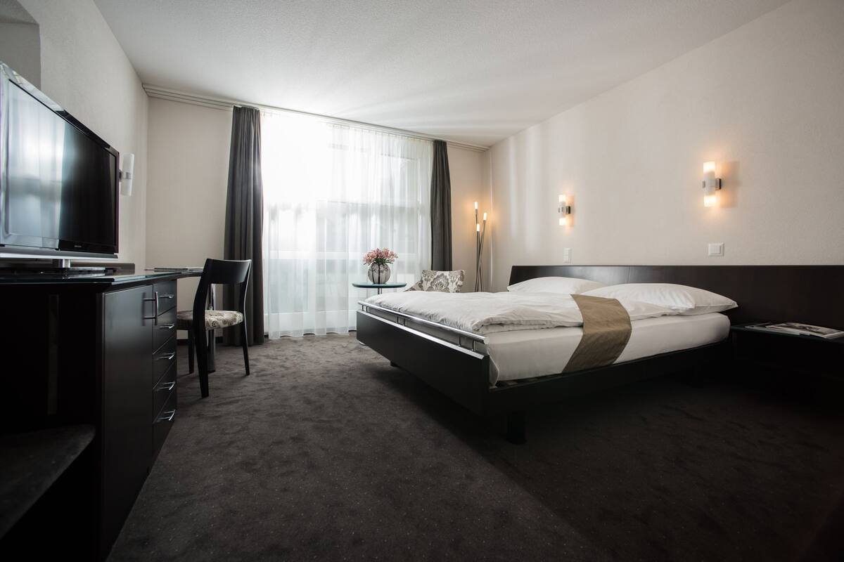 Quarto de hotel com cama, escrivaninha com televisão, uma cadeira e uma mesinha com um vaso de flores.