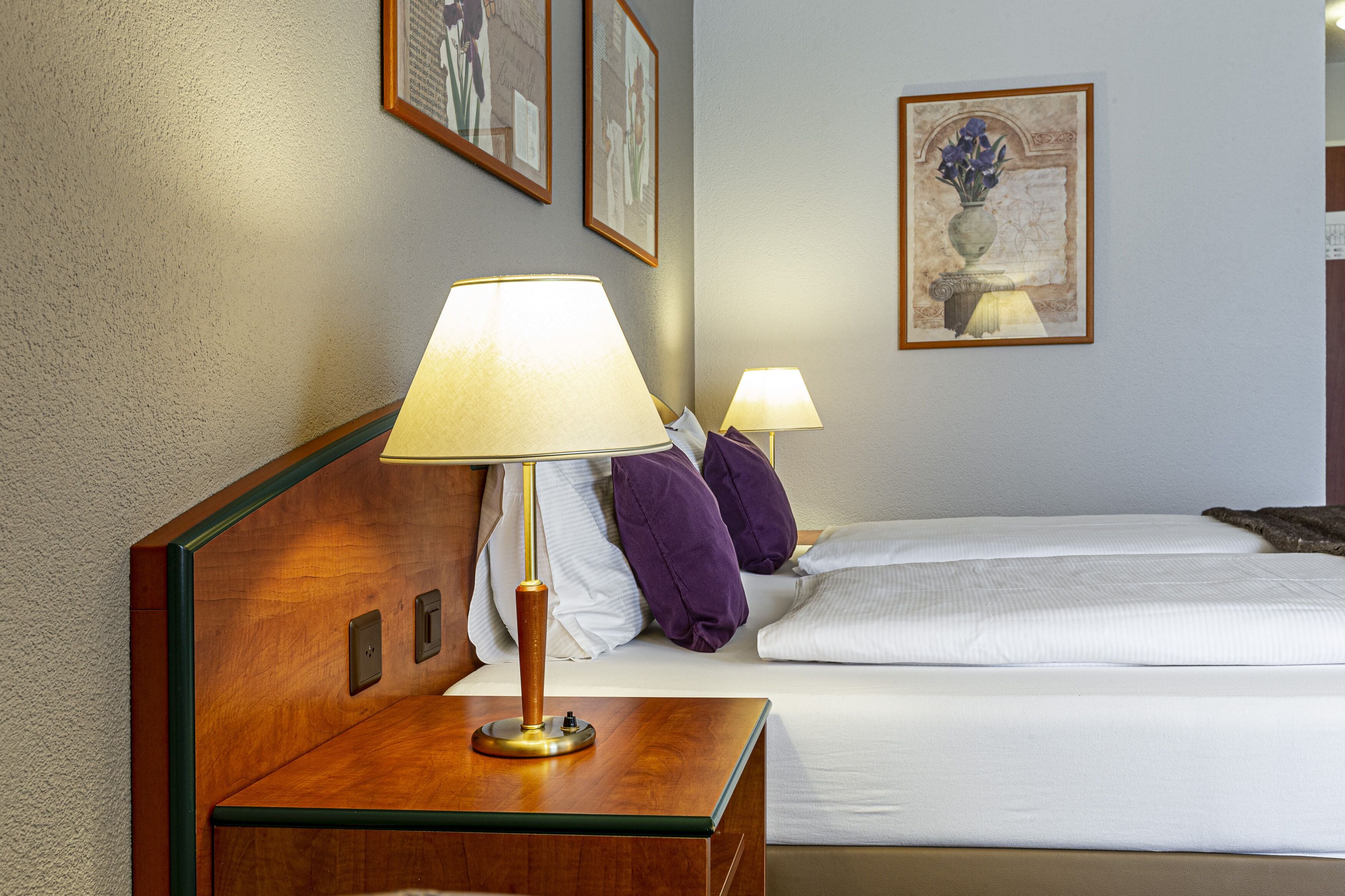 Quarto de hotel com cabeceira de madeira, duas camas com roupa de cama branca, almofadas roxas e duas luminárias de cabeceira.