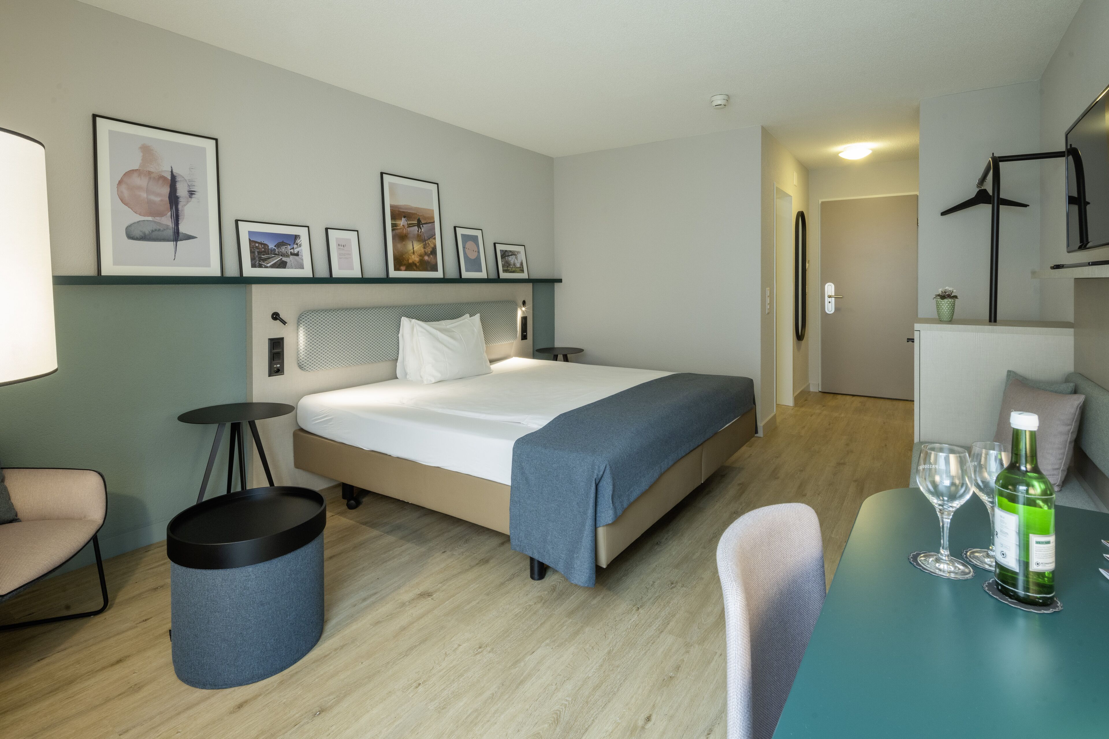 Quarto de hotel moderno com cama, mesa de cabeceira, cadeira e pequena mesa com copos de vinho e uma garrafa.