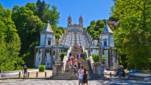 Uma escadaria imponente que leva a um edifício histórico com duas torres, rodeado por vegetação exuberante.