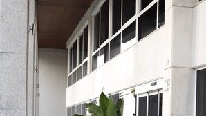Uma área de estar moderna ao ar livre, com cadeiras e mesas brancas, plantas em vasos e um edifício com grandes janelas e piso de azulejos.