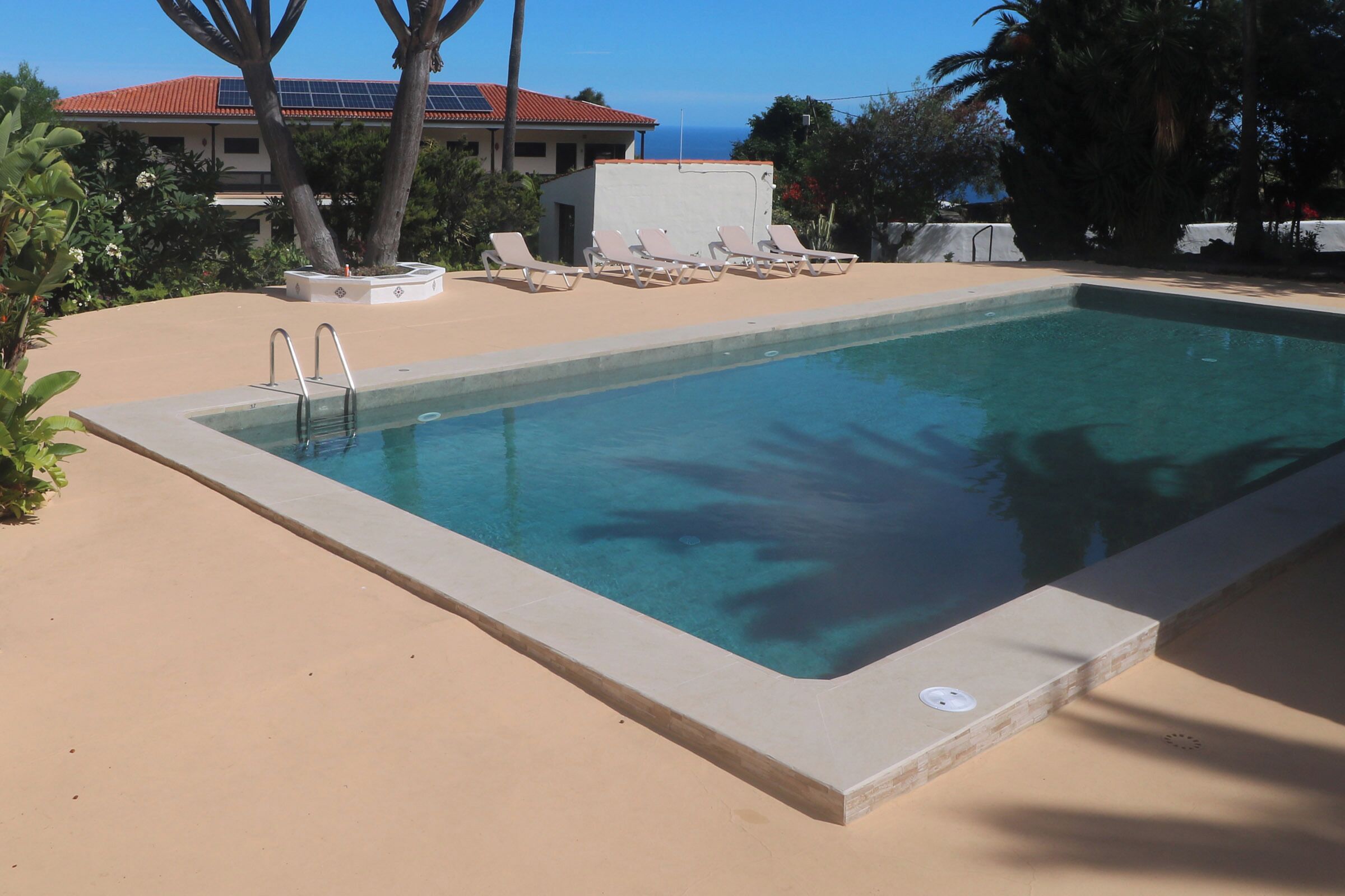 Uma piscina retangular com borda de concreto, rodeada por uma área arenosa com espreguiçadeiras e uma pequena construção.