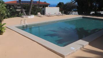 Uma piscina retangular com borda de concreto, rodeada por uma área arenosa com espreguiçadeiras e uma pequena construção.
