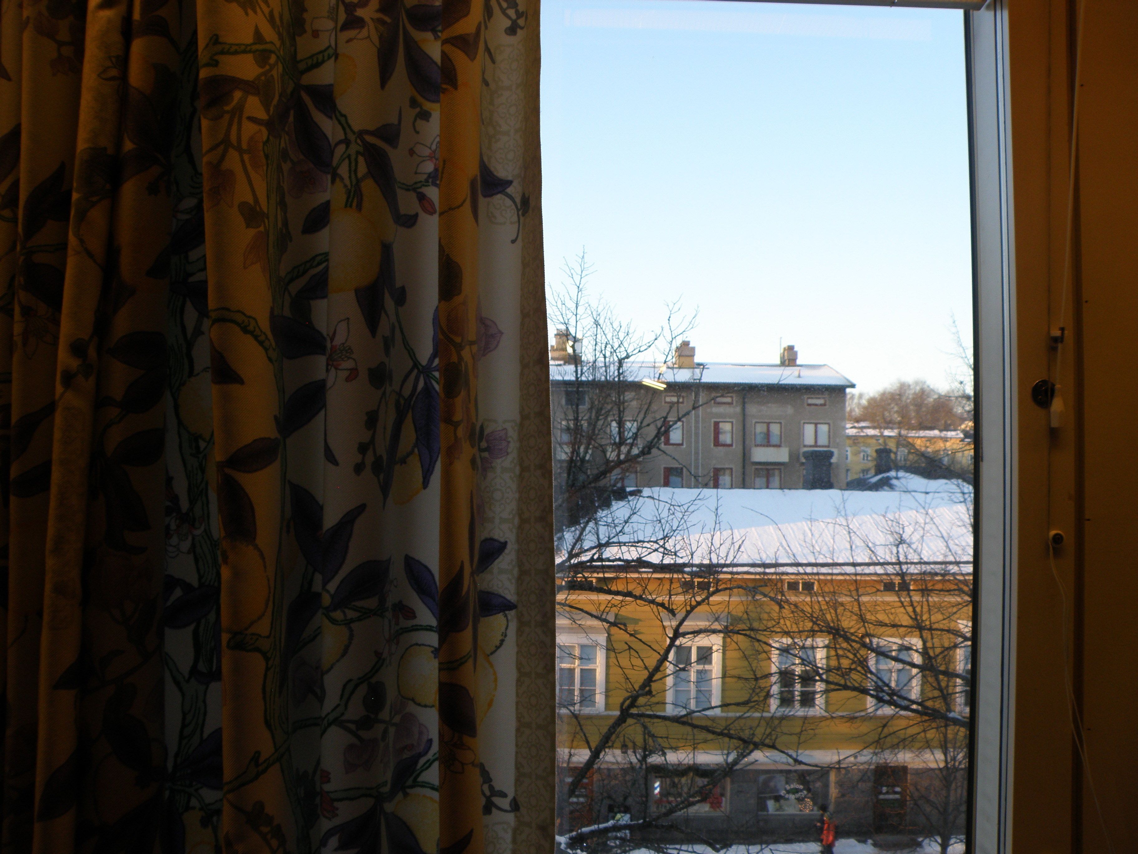 Uma janela com cortinas florais, vista de um prédio com telhado coberto de neve e árvores sem folhas.