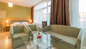 Quarto de hotel moderno com mesa de centro de vidro, dois sofás, cama com roupa de cama branca, mesa de cabeceira com abajur e janelas grandes com cortinas.