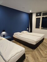 Quarto moderno com duas camas, uma mesa de cabeceira, abajur e uma janela com cortinas.