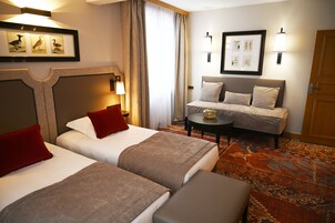 Quarto de hotel com duas camas, uma mesinha e um sofá. Há quadros nas paredes e uma porta à direita.