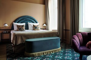 Quarto de hotel com uma cama grande, um banquinho, uma poltrona roxa e um tapete estampado.