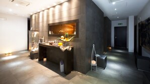 Uma área de recepção moderna com um balcão escuro, uma vitrine de vidro e um pequeno espaço de estar com uma cadeira e um cubo branco.