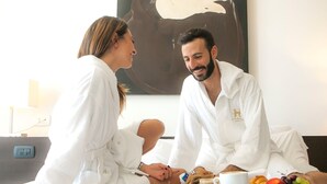 Um casal sentado na cama com café da manhã, incluindo frutas, café e doces.