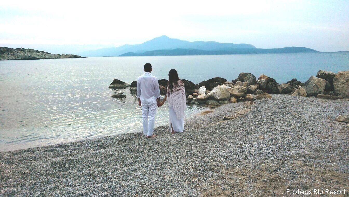 Um casal em pé perto de uma costa rochosa, com vista para um corpo d'água calmo e montanhas ao longe.