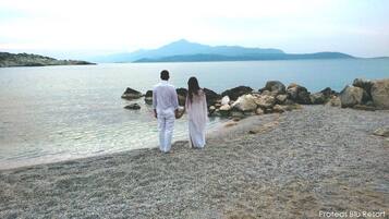 Um casal em pé perto de uma costa rochosa, com vista para um corpo d'água calmo e montanhas ao longe.
