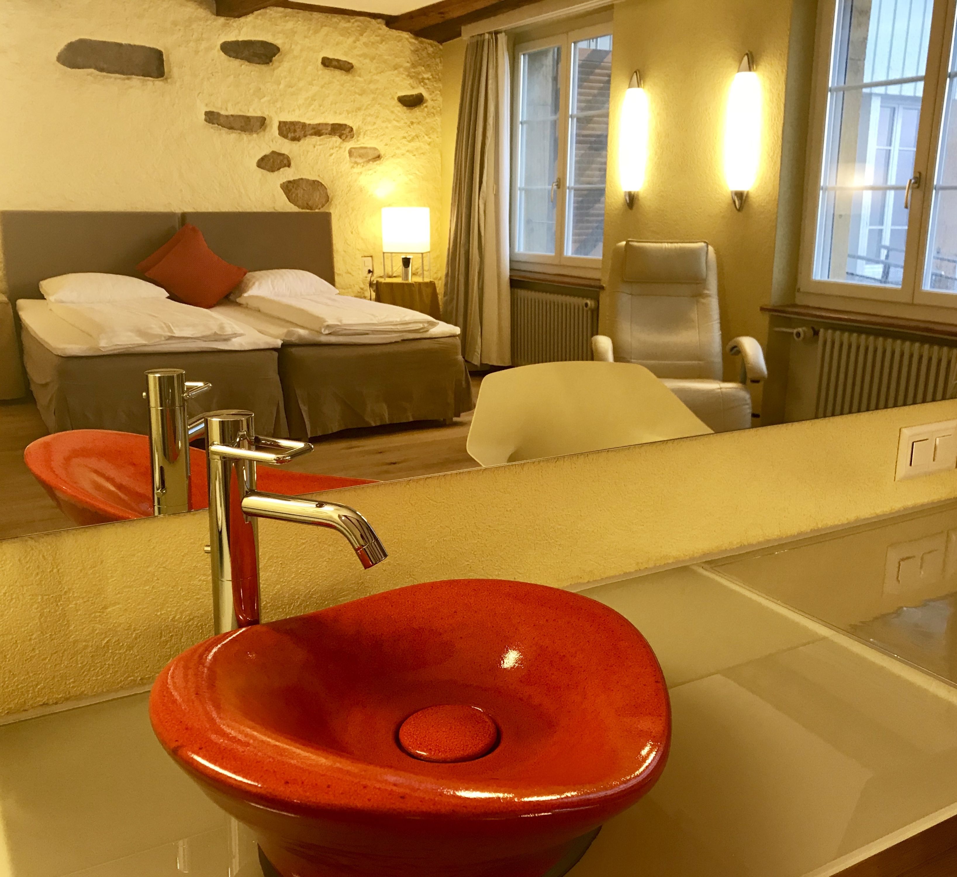 Quarto de hotel com uma pia vermelha, uma cama com roupa de cama branca, uma cadeira e uma parede de pedra.