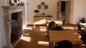 Uma área de refeições com lareira de pedra, mesa de madeira e cadeiras. Há decorações nas paredes e um abajur sobre uma mesinha.