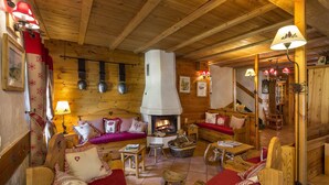 Interior acolhedor de uma cabana de toras, com lareira, móveis de madeira e uma escada que leva a um andar superior.
