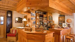 Uma área de bar em madeira com prateleiras de garrafas e copos, um relógio e uma cadeira de madeira com almofada vermelha.