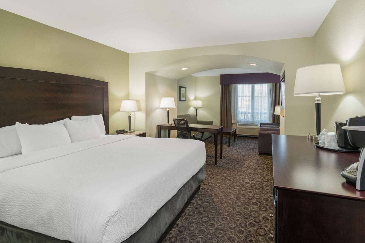 Quarto de hotel com cama grande, escrivaninha, duas cadeiras, luminária e janela com cortinas.