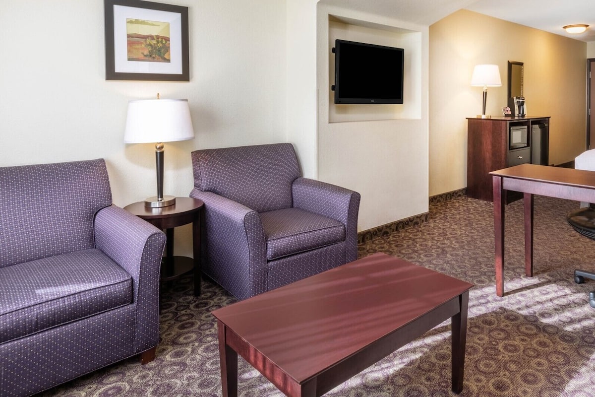 Quarto de hotel com sofá, duas poltronas, escrivaninha, cadeira, televisão e luminária.