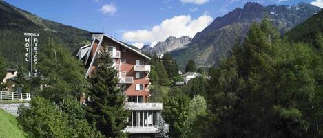 Um hotel com fachada vermelha, várias varandas e vista para montanhas e vegetação.