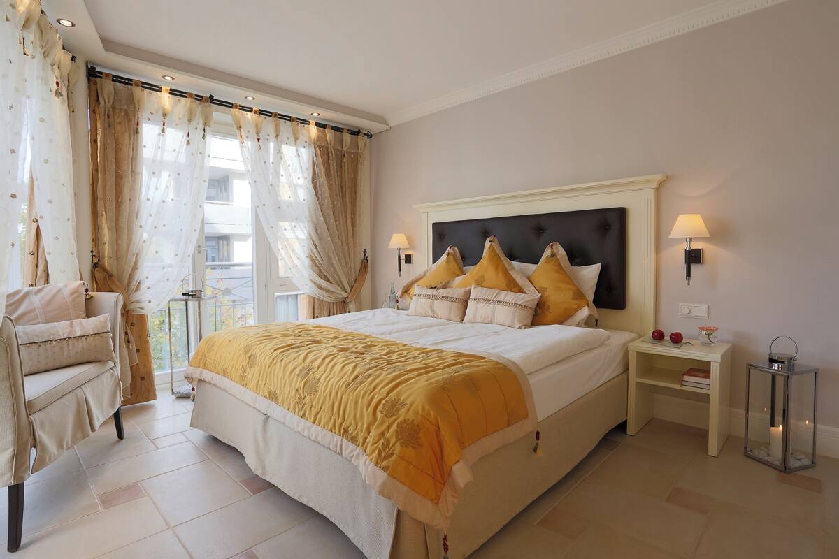 Quarto de hotel com uma cama grande, uma cadeira, uma mesinha de cabeceira com uma luminária e vista para o exterior.