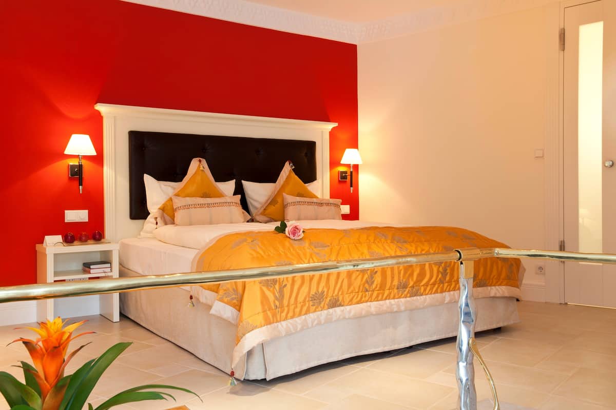 Quarto de hotel com uma parede de destaque vermelha, uma cabeceira branca e uma cama com colcha laranja. Há duas mesas de cabeceira com luminárias e um pequeno estante com livros e objetos decorativos.