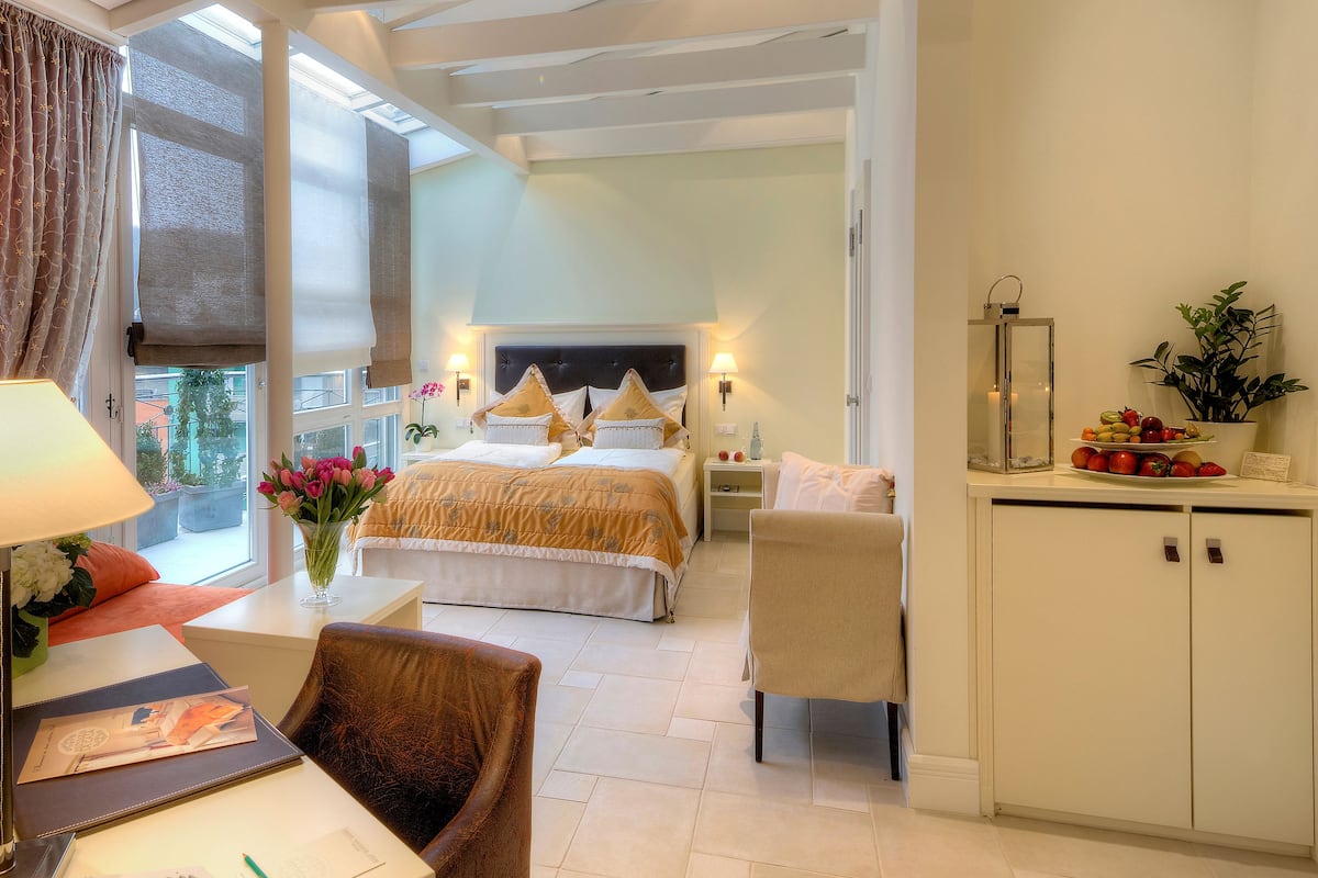 Quarto de hotel moderno com uma cama grande, uma escrivaninha com luminária, uma cadeira, uma mesinha de centro com um vaso de flores e um armário com frutas.