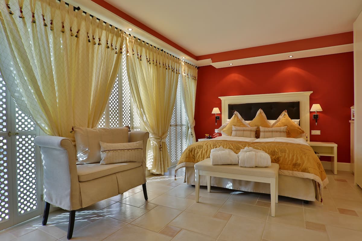 Quarto com cama, mesas de cabeceira, um banco, uma cadeira e cortinas.
