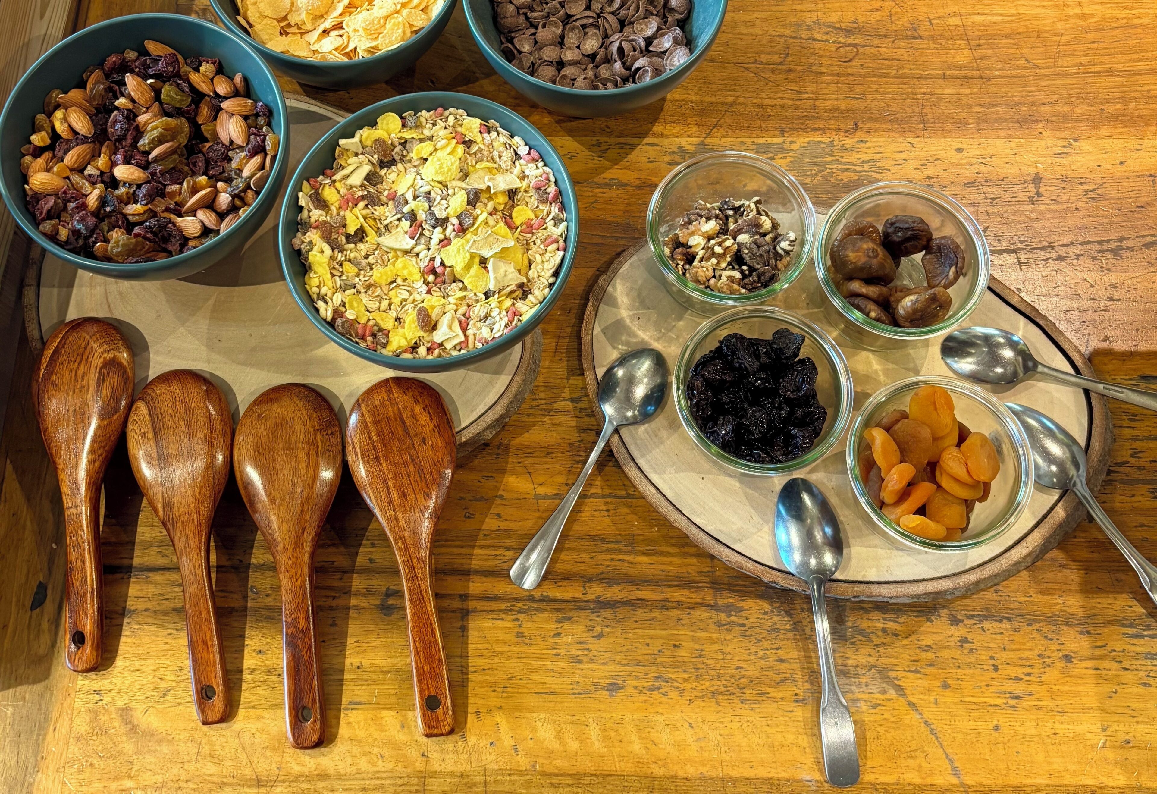 Uma mesa de madeira com tigelas de nozes, frutas secas e granola, acompanhada de colheres de pau e colheres de metal.