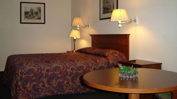 Quarto de hotel com uma cama, duas luminárias fixadas na parede, uma mesa redonda de madeira com um vaso de flores e duas molduras com quadros na parede.