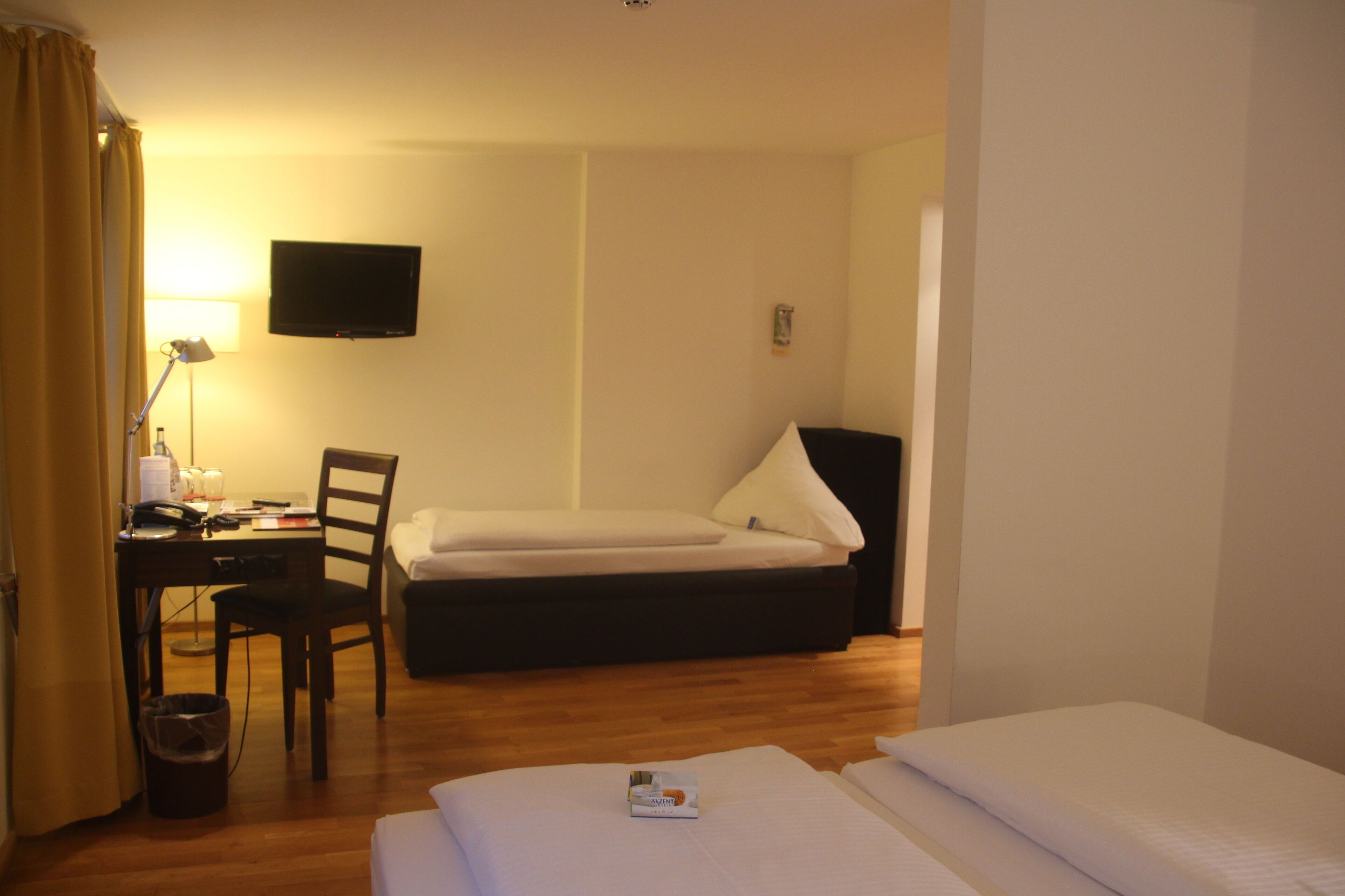 Quarto de hotel com cama, escrivaninha e cadeira, televisão na parede e mesa de cabeceira com abajur.