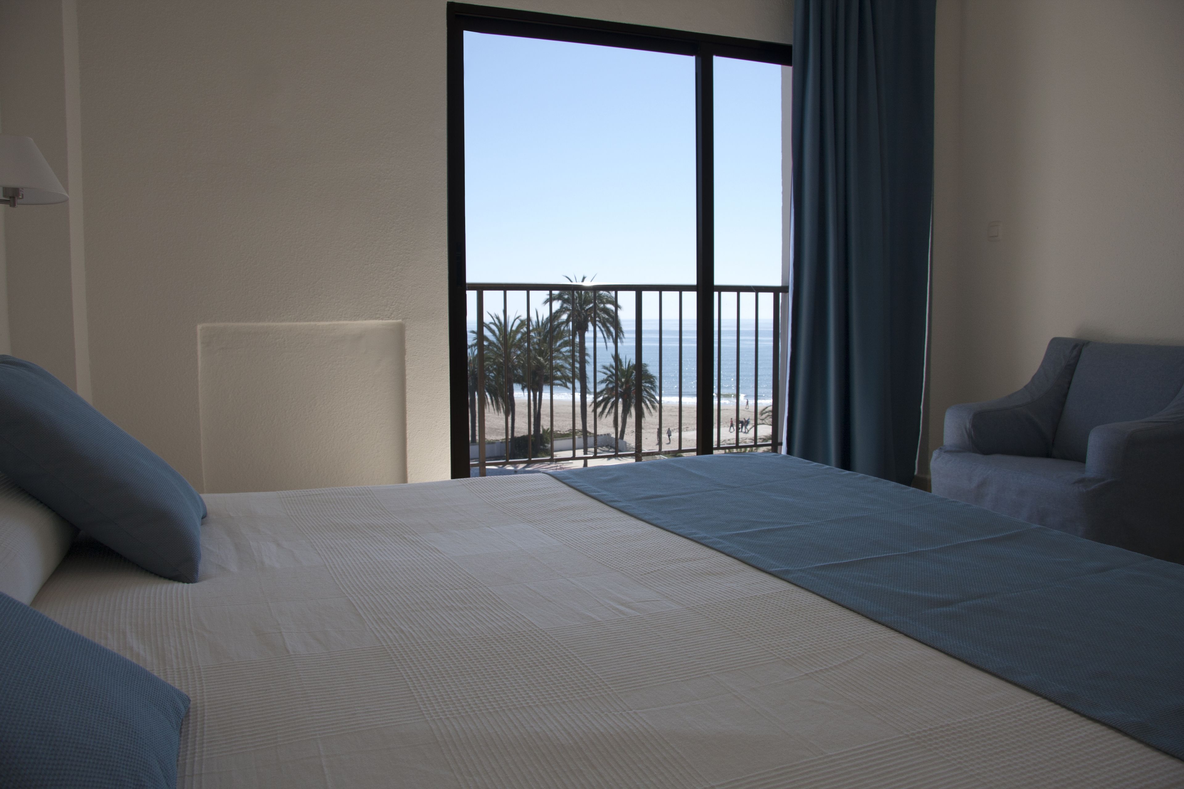 Quarto de hotel com uma cama grande, um sofá e uma varanda com vista para a praia e palmeiras.