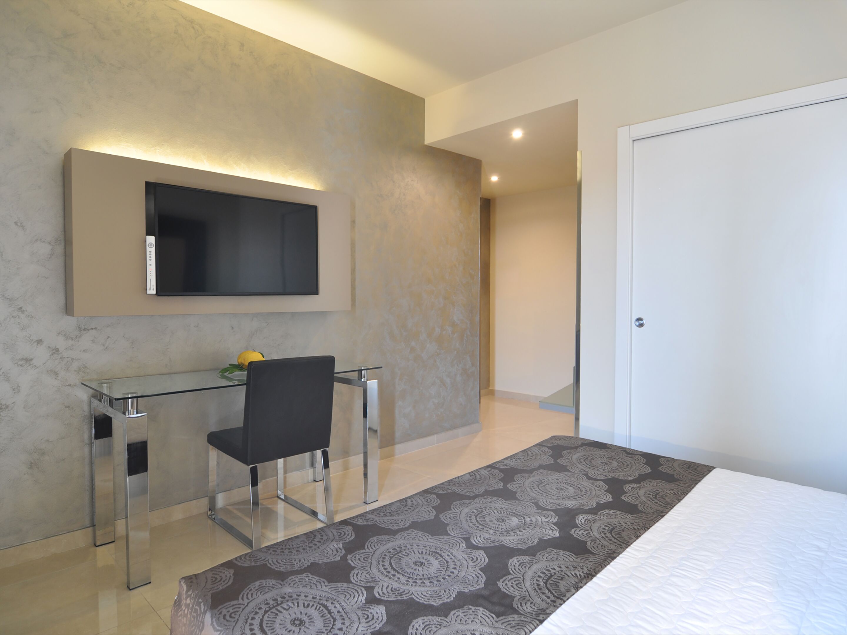 Quarto de hotel moderno com uma cama, uma escrivaninha com cadeira, uma TV de tela plana e uma porta.