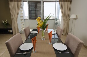 Uma mesa de jantar posta para seis lugares, cadeiras, um vaso com flores e vista da cidade por grandes janelas.