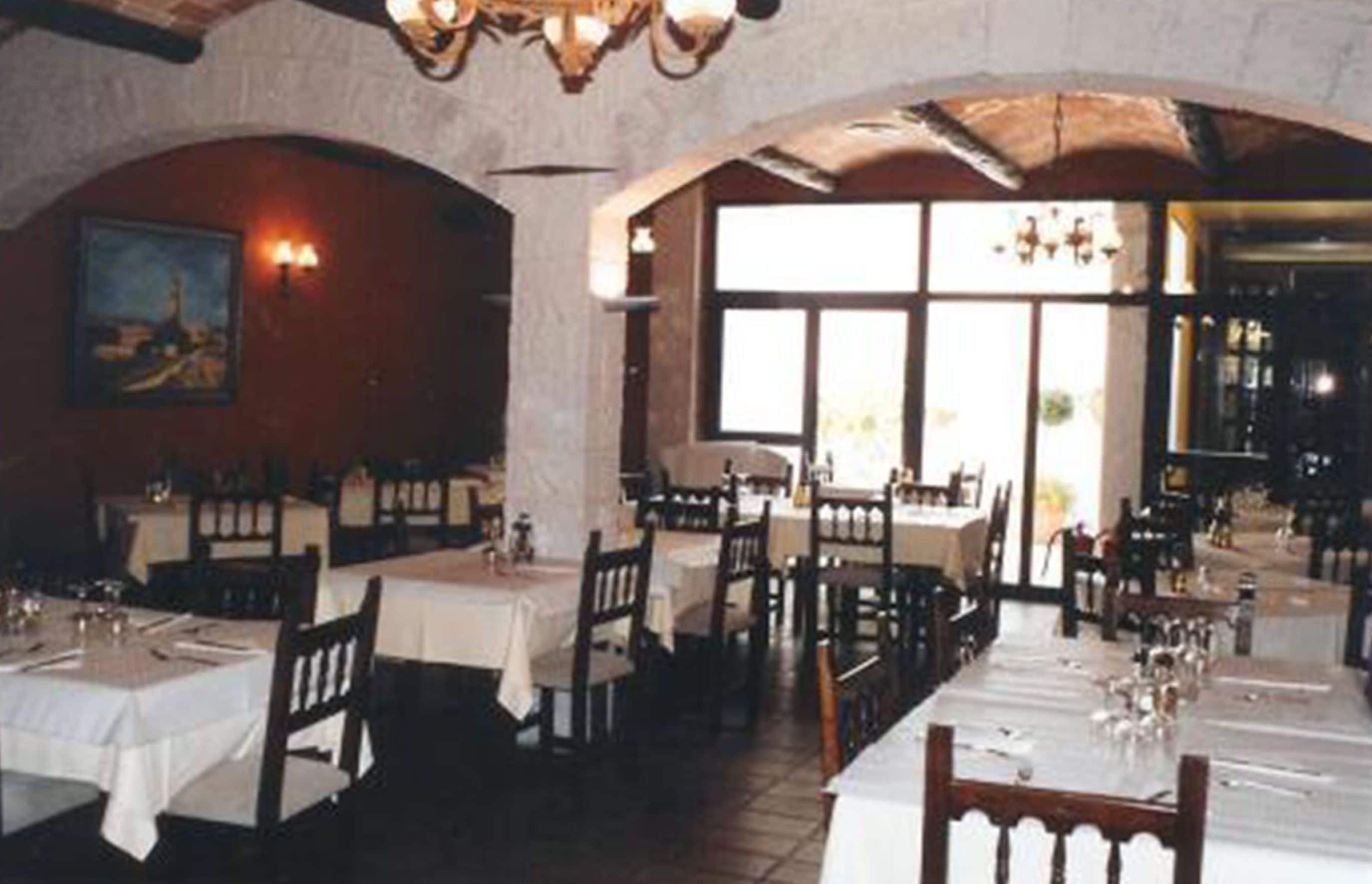 Restaurante com várias mesas e cadeiras, janelas amplas e um candelabro.