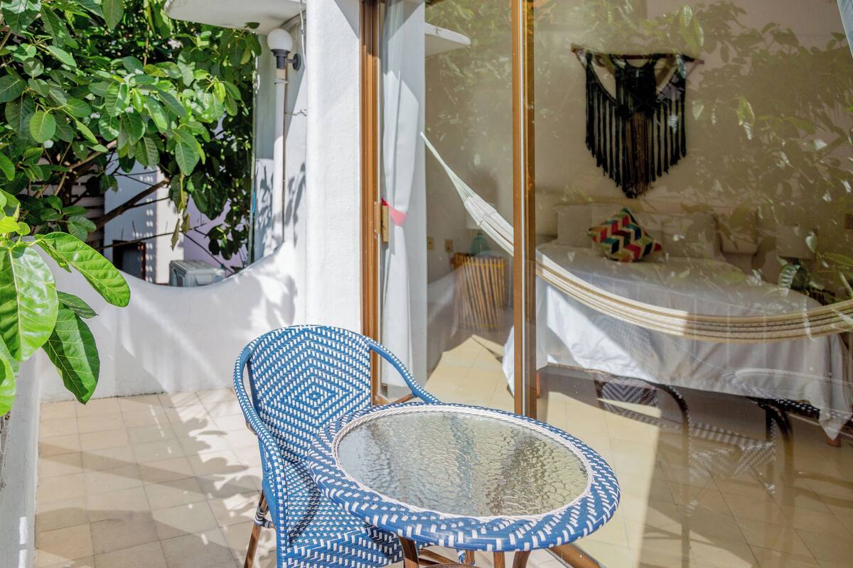 Um pátio ensolarado com uma cadeira azul estampada e uma mesa de vidro, além de uma rede e uma cama com lençol branco.