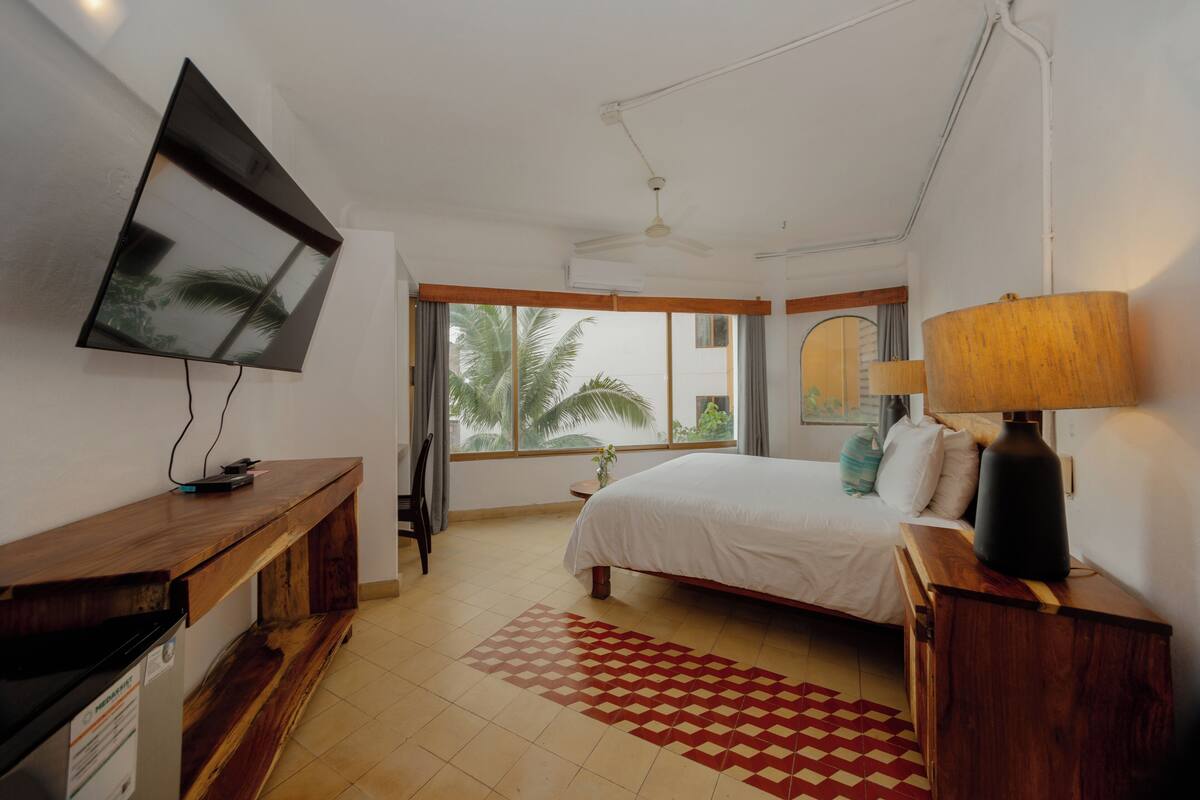 Quarto de hotel com cama, televisão na parede, ventoinha no teto, janela com vista para palmeiras e mesa de cabeceira de madeira com abajur.
